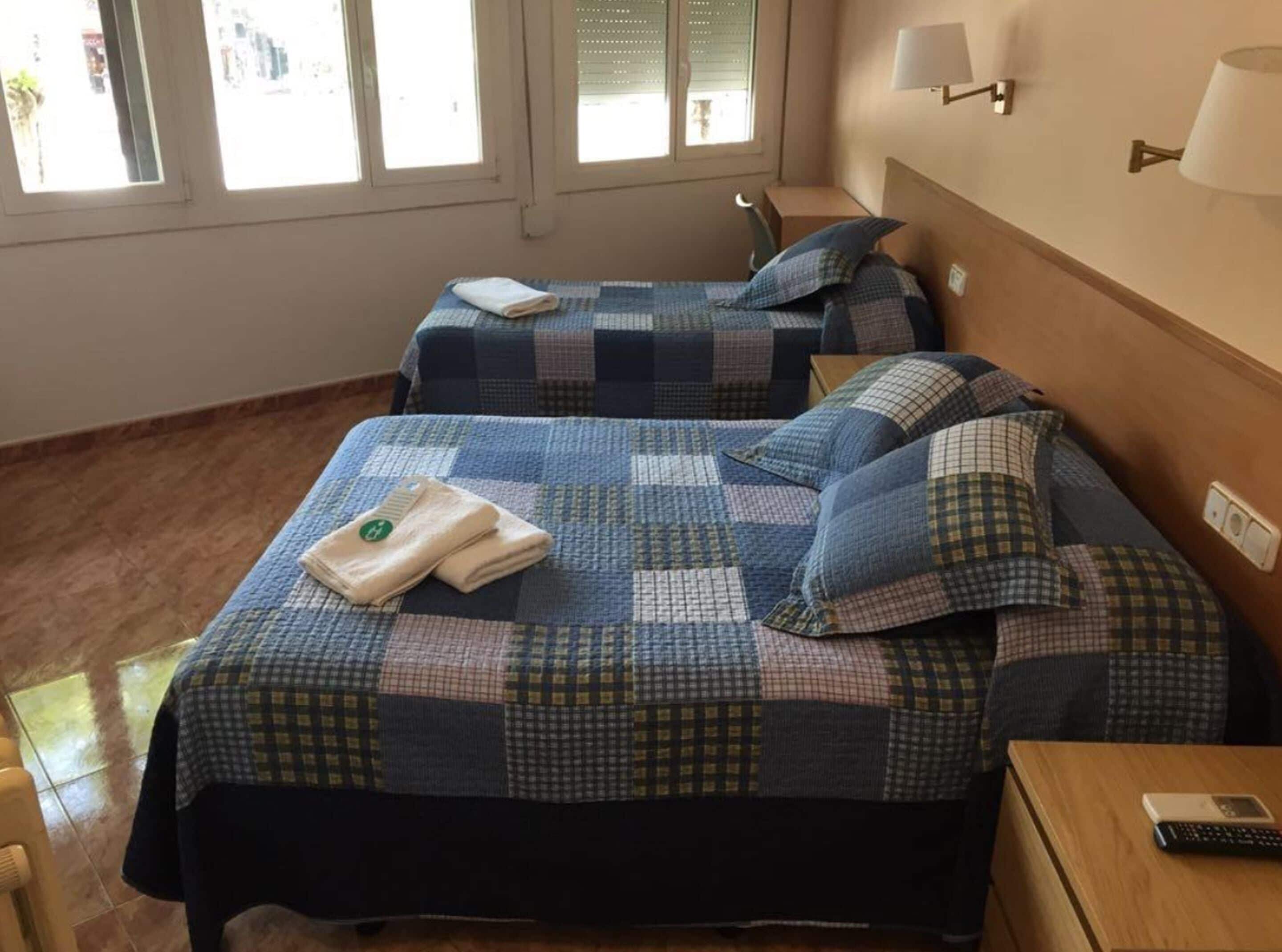 Quarto de hotel com duas camas, cada uma com colcha e almofadas xadrez. Há uma mesa de cabeceira de madeira com uma luminária e um controle remoto.