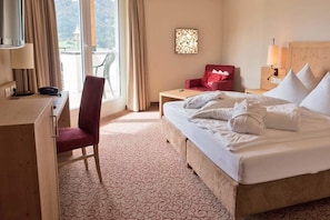 Quarto de hotel com cama, escrivaninha, cadeira, mesa pequena e janela com cortinas.