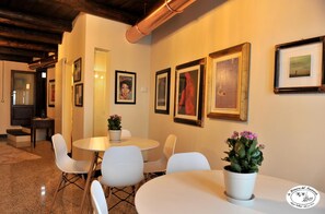 Uma sala de jantar com cadeiras e mesas brancas, obras de arte nas paredes e uma planta em vaso sobre a mesa.