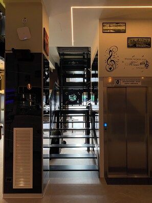 Um saguão de hotel com uma escadaria que leva à área de recepção, um sinal indicando a localização da recepção e do bar, e uma parede com design de nota musical e texto.