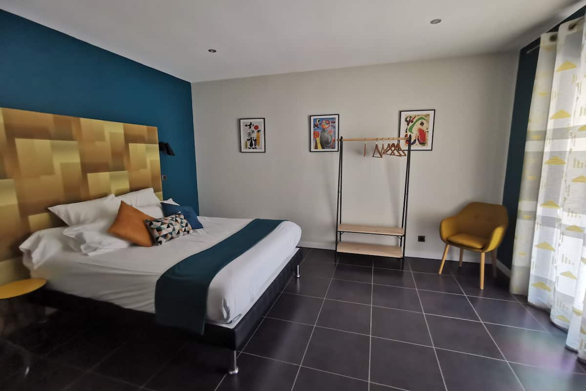 Quarto de hotel moderno com uma cama grande, cabeceira de madeira, uma cadeira pequena e obras de arte nas paredes.