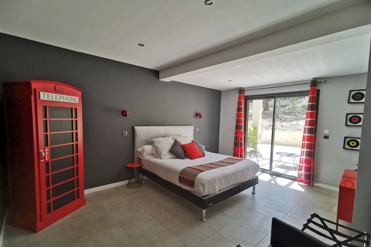 Quarto moderno com cama, uma cabine telefônica vermelha e vista para o exterior.
