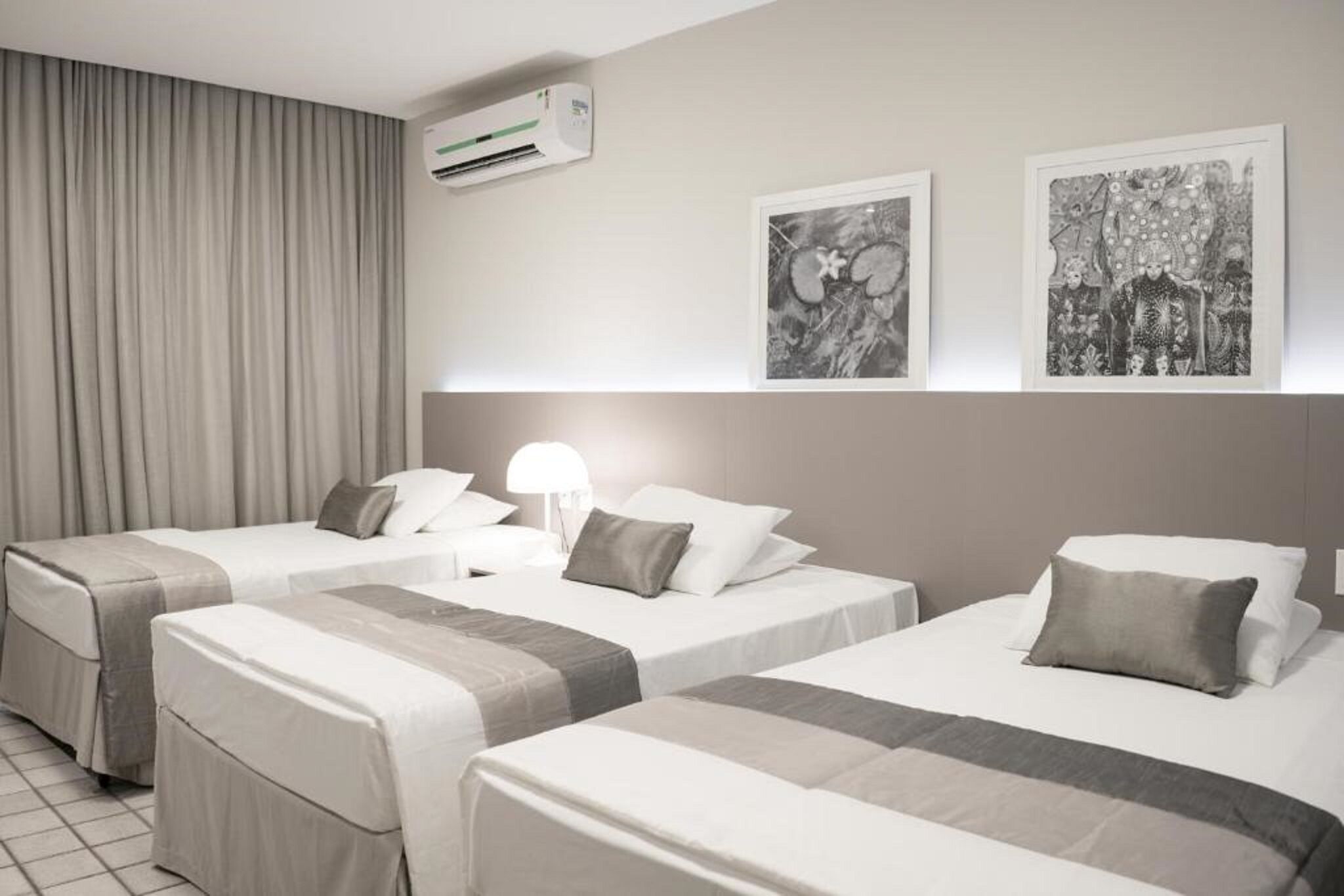 Quarto de hotel com três camas, uma janela grande com cortinas, ar condicionado e duas obras de arte emolduradas na parede.