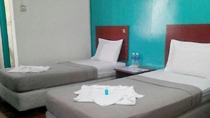 Quarto de hotel com duas camas de solteiro, cada uma com colchão cinzento e roupa de cama branca. Há uma pequena mesa com uma garrafa de água e um bloco de notas entre as camas.