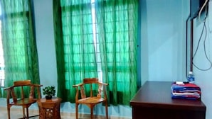 Quarto com duas cadeiras de madeira, uma mesinha com uma planta, uma escrivaninha com garrafa de água e roupas dobradas, e cortinas verdes.