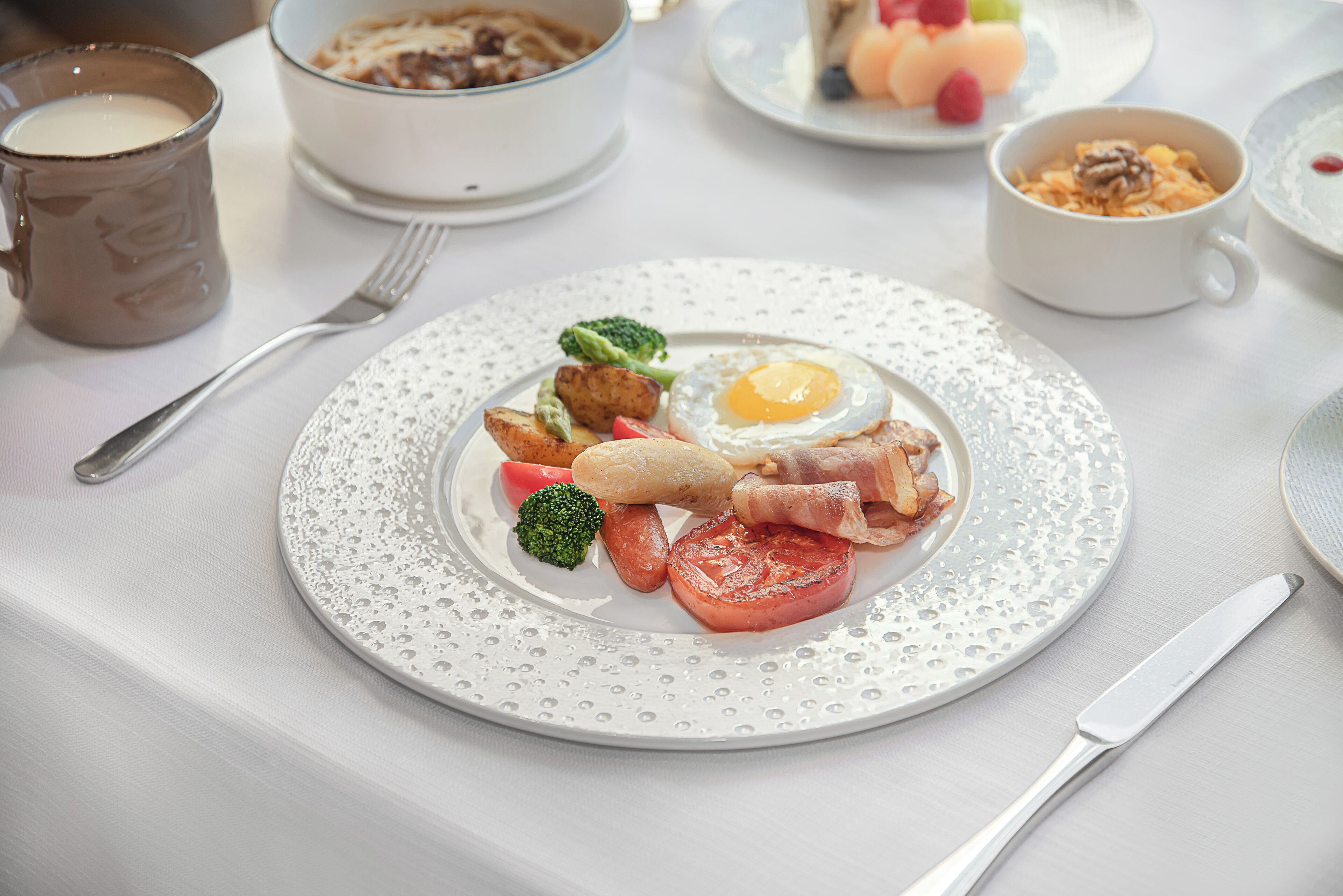 Prato de pequeno-almoço com ovos, bacon, tomate, brócolis e salsicha, sobre um prato branco com padrão texturizado.