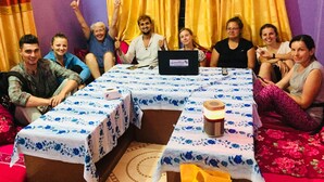 Um grupo de pessoas sentadas à mesa com um laptop e uma xícara, em um quarto com cortinas roxas e amarelas.