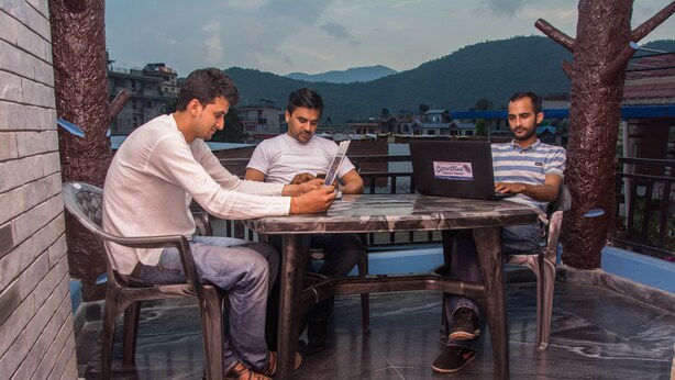 Três homens sentados à volta de uma mesa com um laptop, num espaço ao ar livre com montanhas ao fundo.