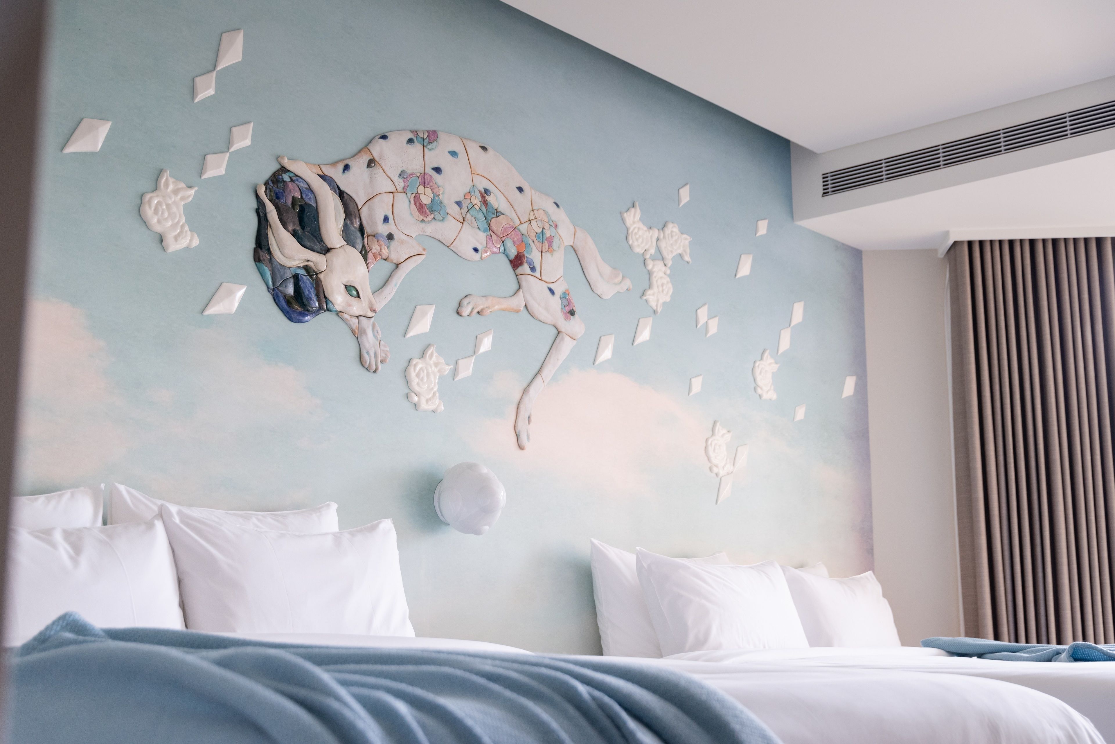 Quarto de hotel com uma cama, almofadas brancas, manta azul, painel na parede com pintura de um cavalo e uma janela com cortinas.
