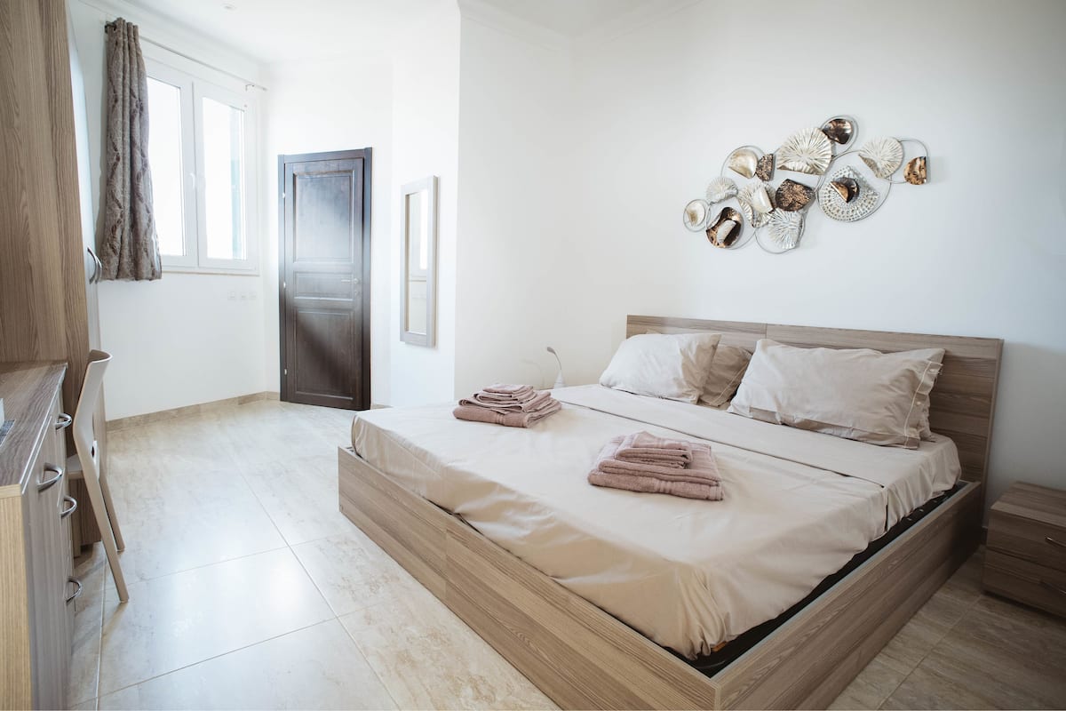 Quarto com cama de madeira, mesa de cabeceira, uma obra de arte na parede e janela com cortinas.