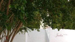 Uma área externa com mesas e cadeiras redondas, rodeada por um muro branco e uma árvore que oferece sombra.