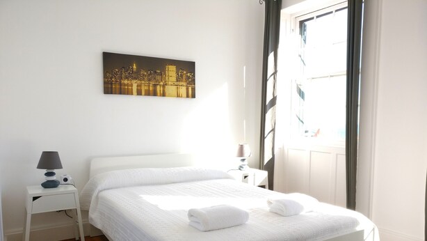 Cama bem arrumada com lençóis brancos e duas toalhas, uma pequena mesa de cabeceira com abajur, um quadro com uma vista urbana na parede e uma janela com cortinas.