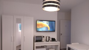 Quarto compacto com escrivaninha branca, televisão montada acima, micro-ondas, mini-geladeira, mesa branca e luminária de teto listrada.