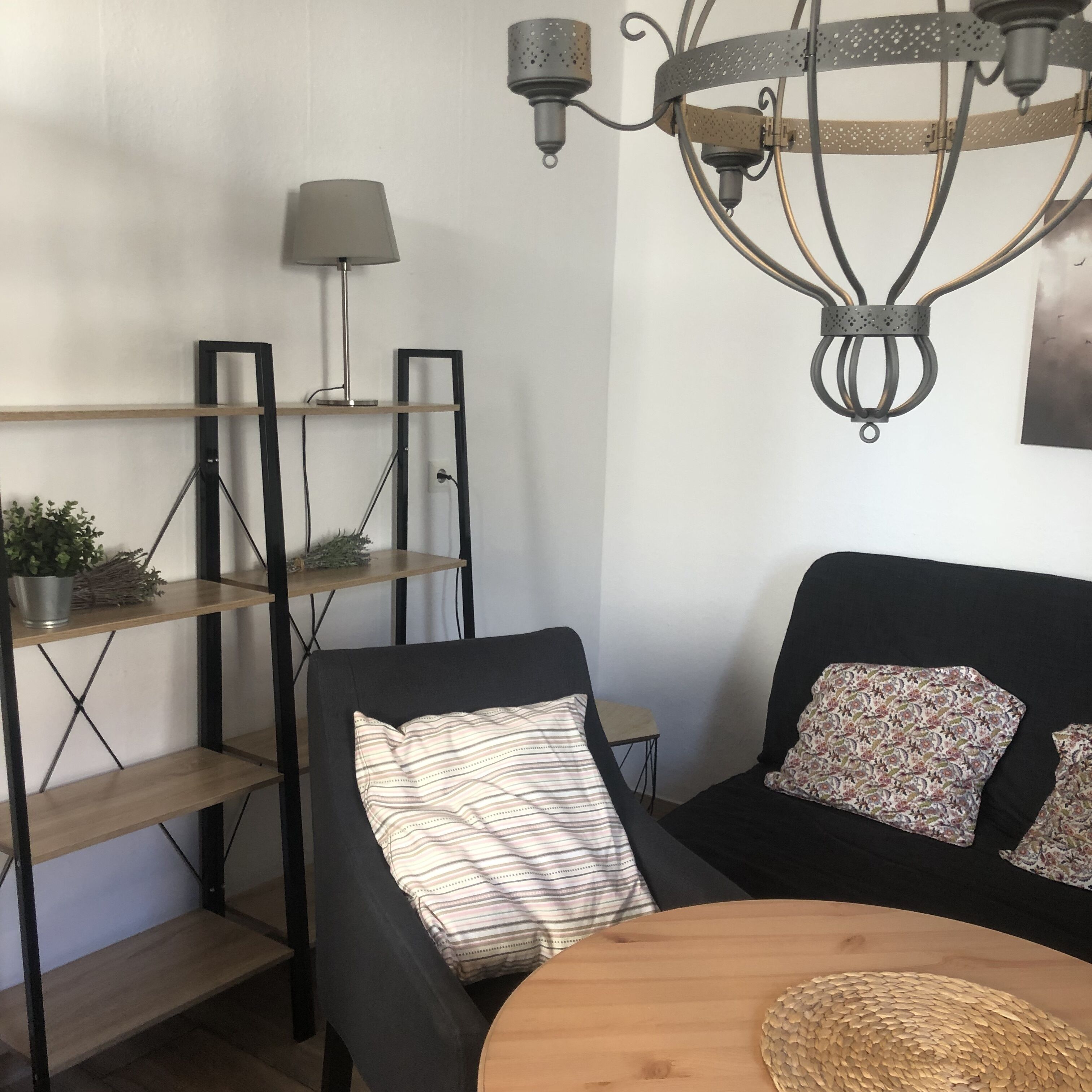 Uma sala de estar moderna com um sofá preto, almofadas listradas, uma mesa redonda de madeira e prateleiras de metal com um abajur e uma pequena planta.