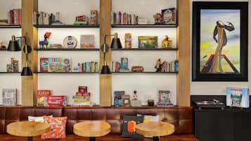Um espaço acolhedor para leitura, com poltronas de couro, mesas redondas e uma estante cheia de livros e jogos de tabuleiro.