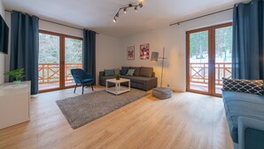 Ampla sala de estar com sofá, poltrona, mesa de centro e um rack para TV. Grandes janelas proporcionam vista para um deck de madeira e árvores.