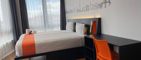 Quarto de hotel com uma cama, uma escrivaninha e uma cadeira. Há uma janela com cortinas e uma parede com um mural da paisagem urbana.