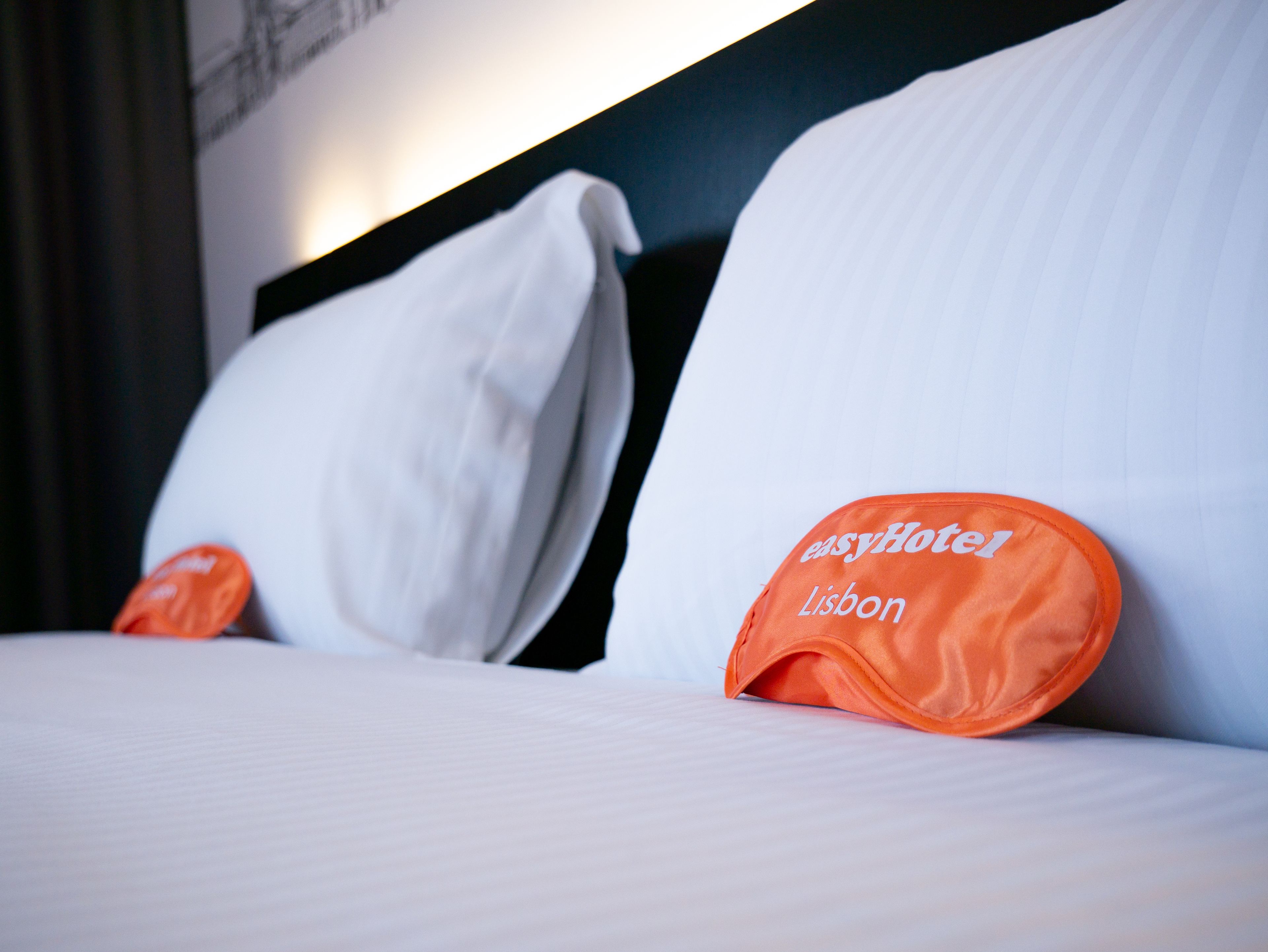Uma cama de hotel com almofadas brancas e uma almofada laranja com o texto 'easyHotel Lisboa'.