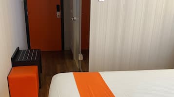 Quarto de hotel com uma cama, um banquinho laranja pequeno, um controle remoto e uma toalha enrolada.
