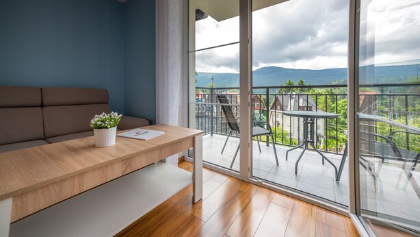 Quarto moderno com um banco de madeira, uma pequena mesa e uma planta em vaso. Possui grandes portas de vidro que dão para uma varanda externa com mesa e cadeiras.