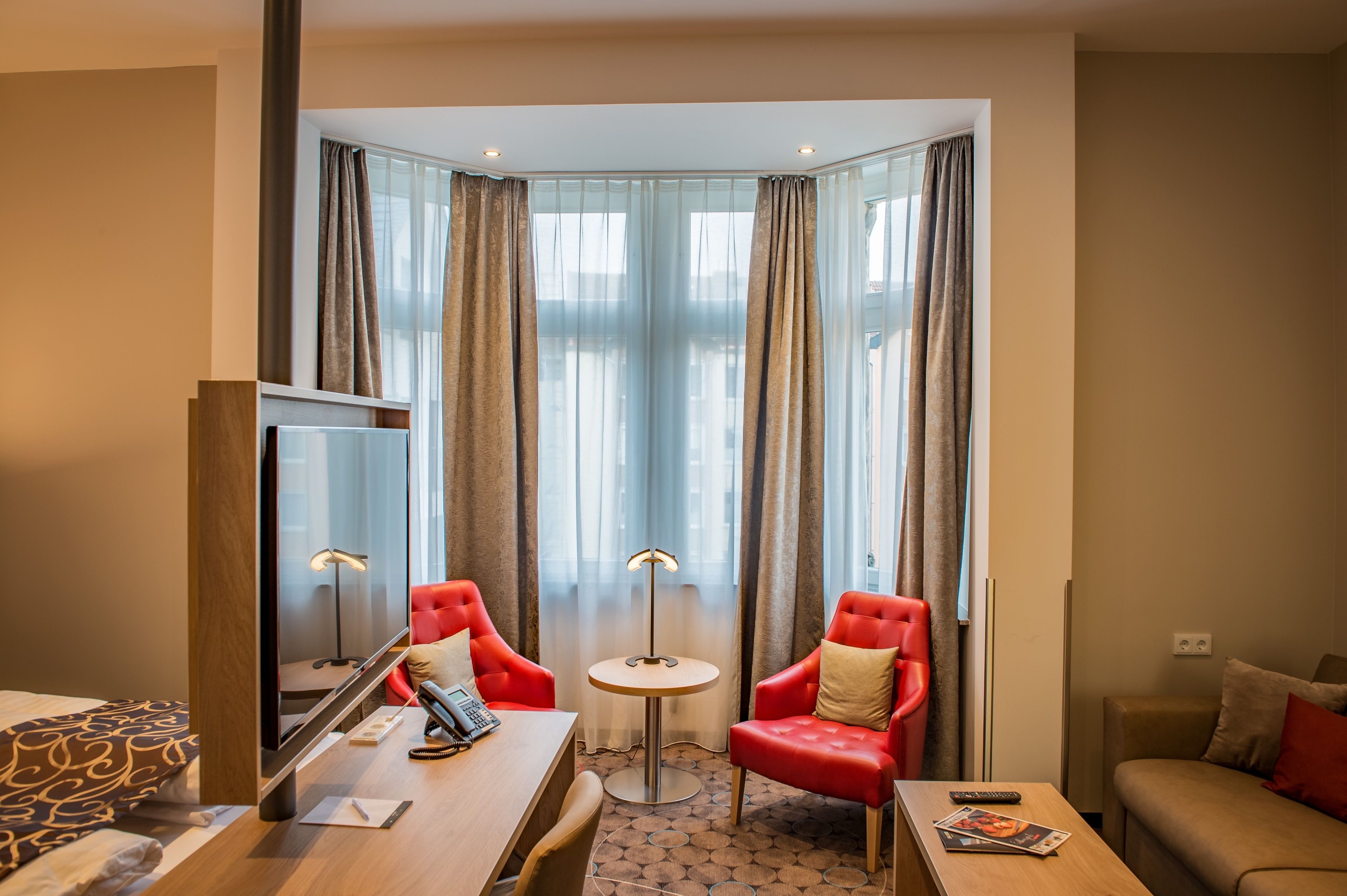 Quarto de hotel com cama, escrivaninha com luminária, duas cadeiras vermelhas, mesa redonda pequena e um sofá.