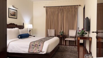 Quarto de hotel com uma cama grande, duas cadeiras, uma mesinha com um vaso e uma televisão.