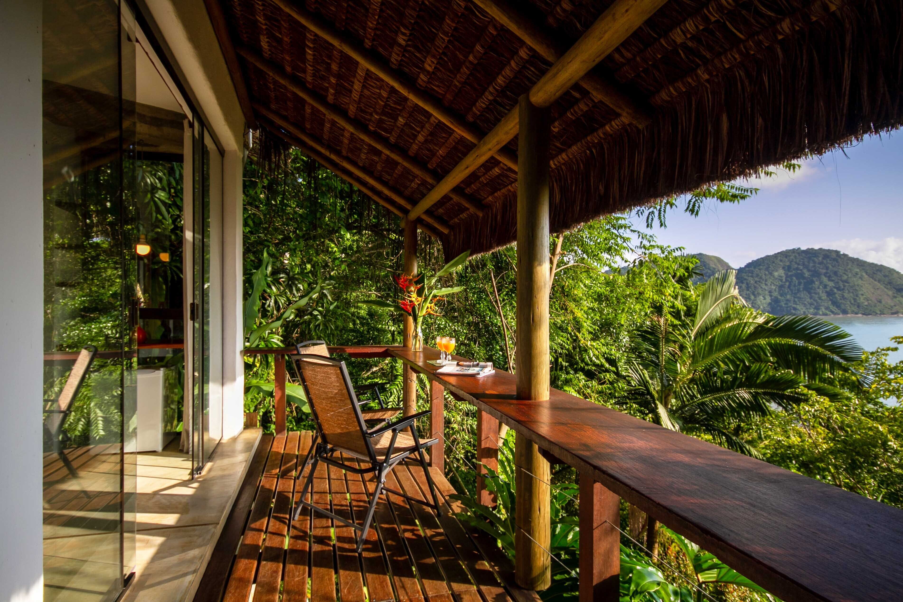 Um deck de madeira com guarda-corpo de vidro, com vista para vegetação exuberante e um corpo de água.