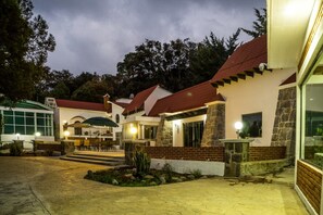 Um pátio bem iluminado com um caminho de pedra, uma área de refeições com mesas ao ar livre e um edifício com telhado vermelho e paredes de pedra.