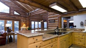 Interior de uma cabana rústica com uma grande ilha na cozinha, armários de madeira e vista pelas janelas.