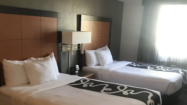 Quarto de hotel com duas camas, cabeceira, mesa de cabeceira com abajur e janela com cortinas.