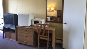 Quarto de hotel compacto com escrivaninha de madeira, televisão, micro-ondas, mini-geladeira, cadeira de madeira e espelho refletindo um abajur e uma cama.
