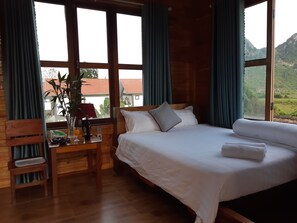 Quarto com estrutura de madeira, cama, mesa com garrafa e copos, e vista para uma paisagem de montanhas.