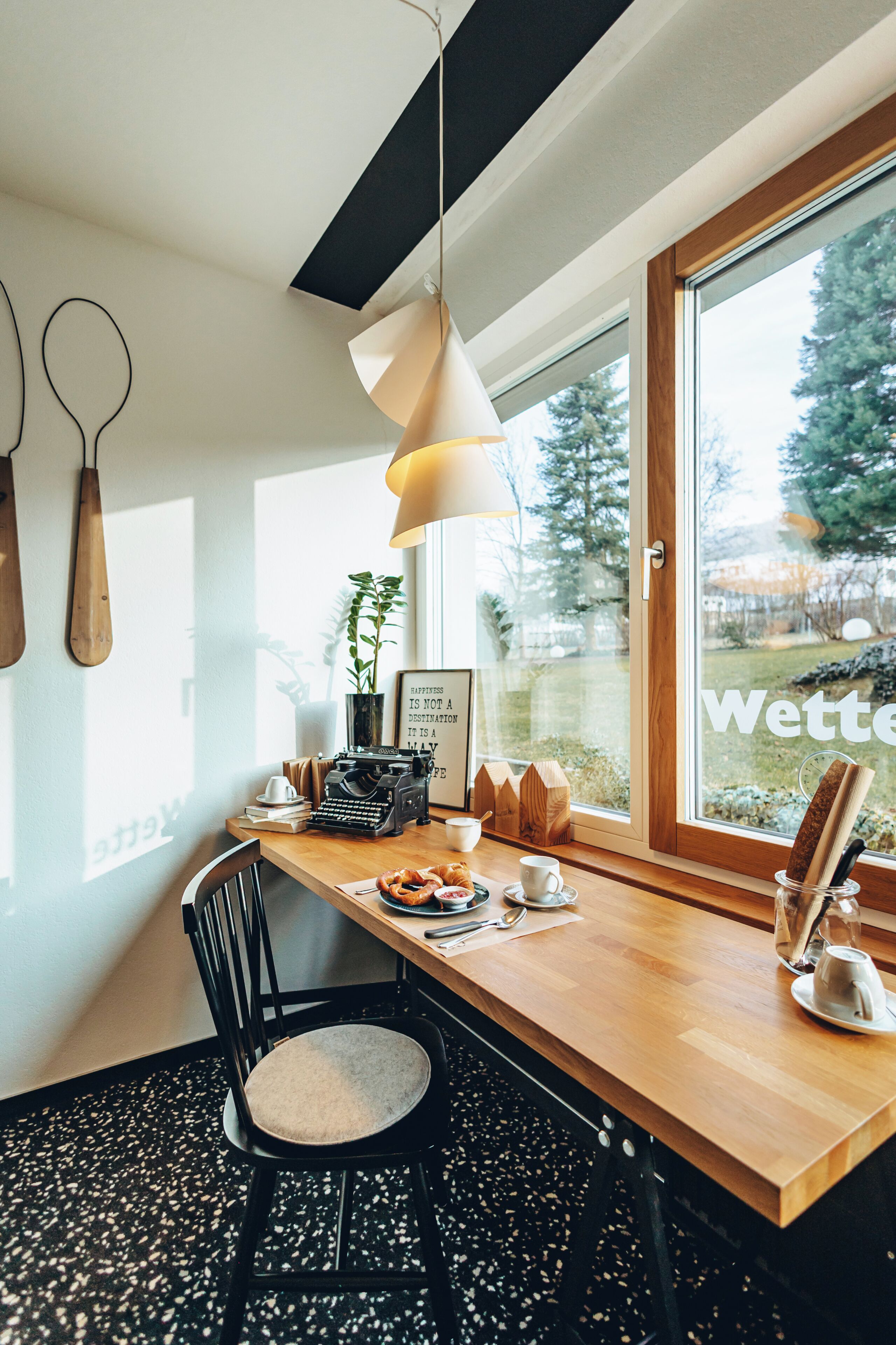 Uma zona de refeições moderna com uma mesa de madeira, uma cadeira preta, um candeeiro pendente e uma janela com vista para vegetação.