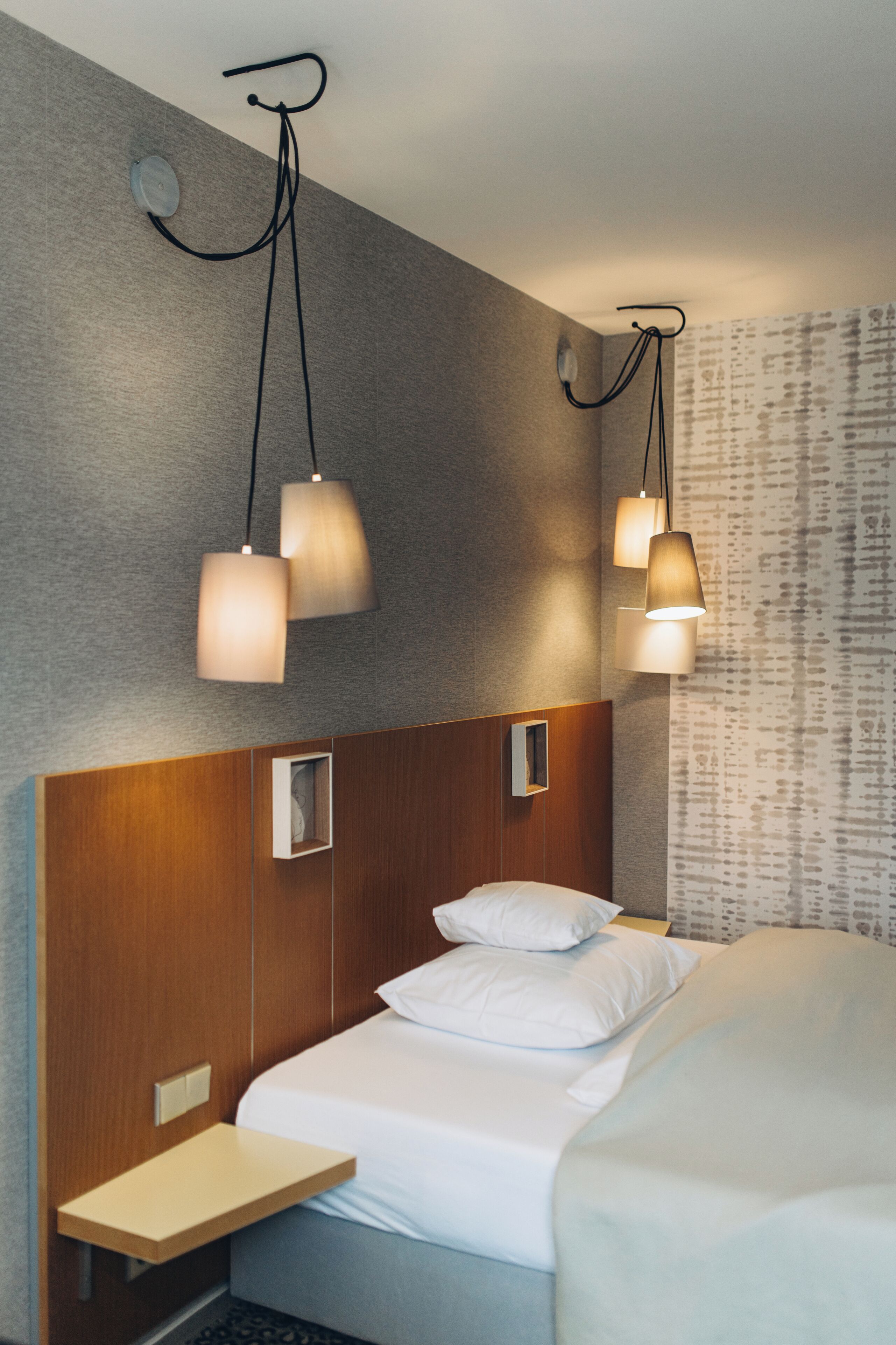 Quarto de hotel com cabeceira de madeira, mesa de cabeceira, cama com roupa de cama branca e duas luminárias fixadas na parede.