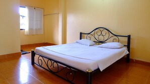 Uma cama de solteiro com estrutura de metal e roupa de cama branca, num quarto com paredes amarelas e soalho de madeira.