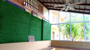 Quarto com parede de grama sintética verde, bancos de madeira, ventilador de teto e janela com vista para palmeiras.