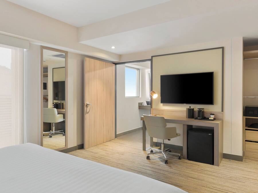 Quarto de hotel com cama, escrivaninha, televisão, guarda-roupa e um pequeno frigorífico.