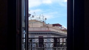 Vista de dentro de um edifício, através de portas abertas, para uma varanda com guarda-corpo de metal, piso de azulejos e edifícios ao fundo.