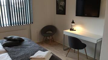 Quarto com cama, escrivaninha, cadeira, luminária, televisão e janela com persianas.