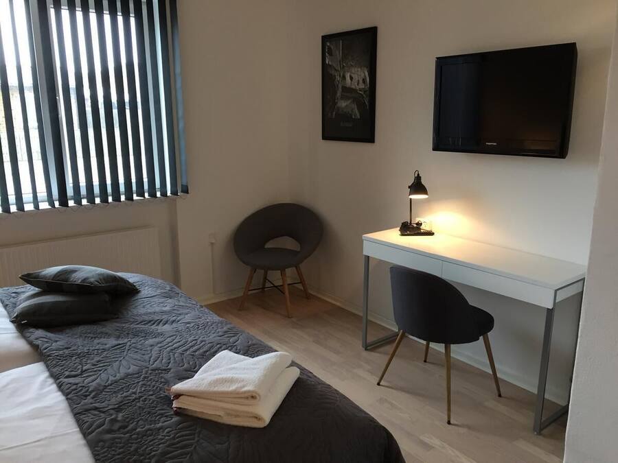 Quarto com cama, escrivaninha, cadeira, luminária, televisão e janela com persianas.