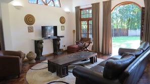 Uma sala de estar ampla com um sofá de couro, uma mesa de centro de madeira e uma televisão. Há objetos decorativos nas paredes e uma grande janela que oferece iluminação natural.