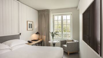 Quarto de hotel com cama, escrivaninha com luminária, cadeira, mesinha com planta e uma grande janela com vista para árvores.