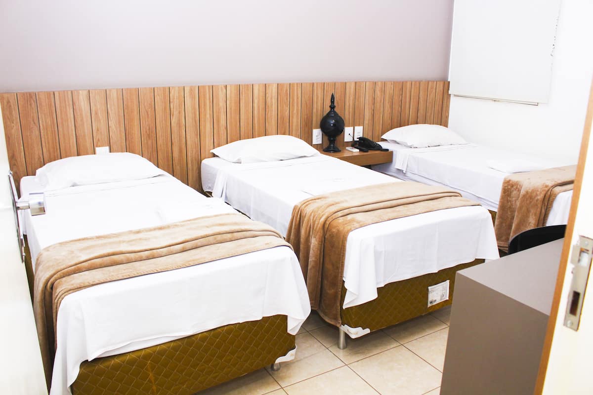 Quarto de hotel com três camas, painel de cabeceira em madeira, roupa de cama branca e mesa de cabeceira com abajur e telefone.