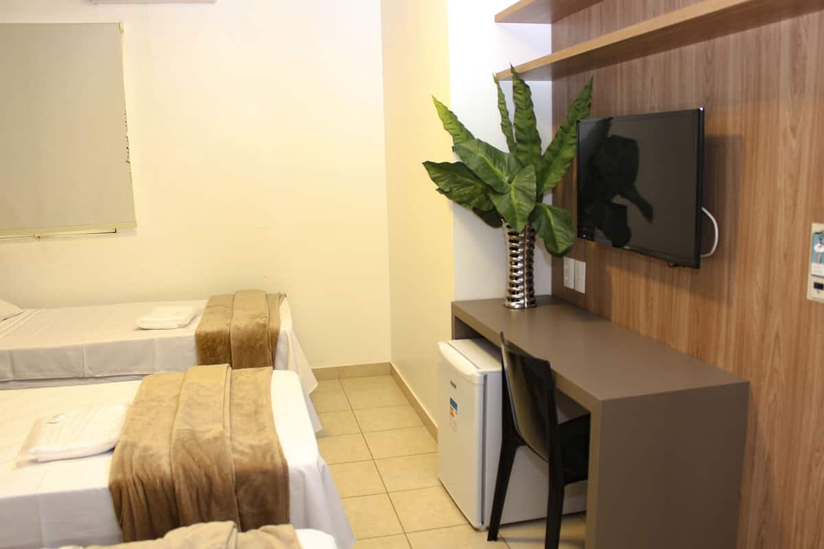 Quarto de hotel com duas camas, uma escrivaninha, televisão, mini-geladeira e um vaso de planta.