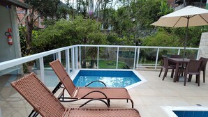 Uma área à beira da piscina com espreguiçadeiras de vime, uma mesa com cadeiras e um grande guarda-sol.