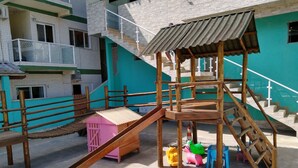 Um parquinho com um escorrega de madeira e estruturas de escalada, cercado por um edifício com fachada verde e várias janelas.
