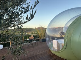 Estrutura em cúpula transparente com deck de madeira, mobiliário ao ar livre e vista para uma paisagem montanhosa.