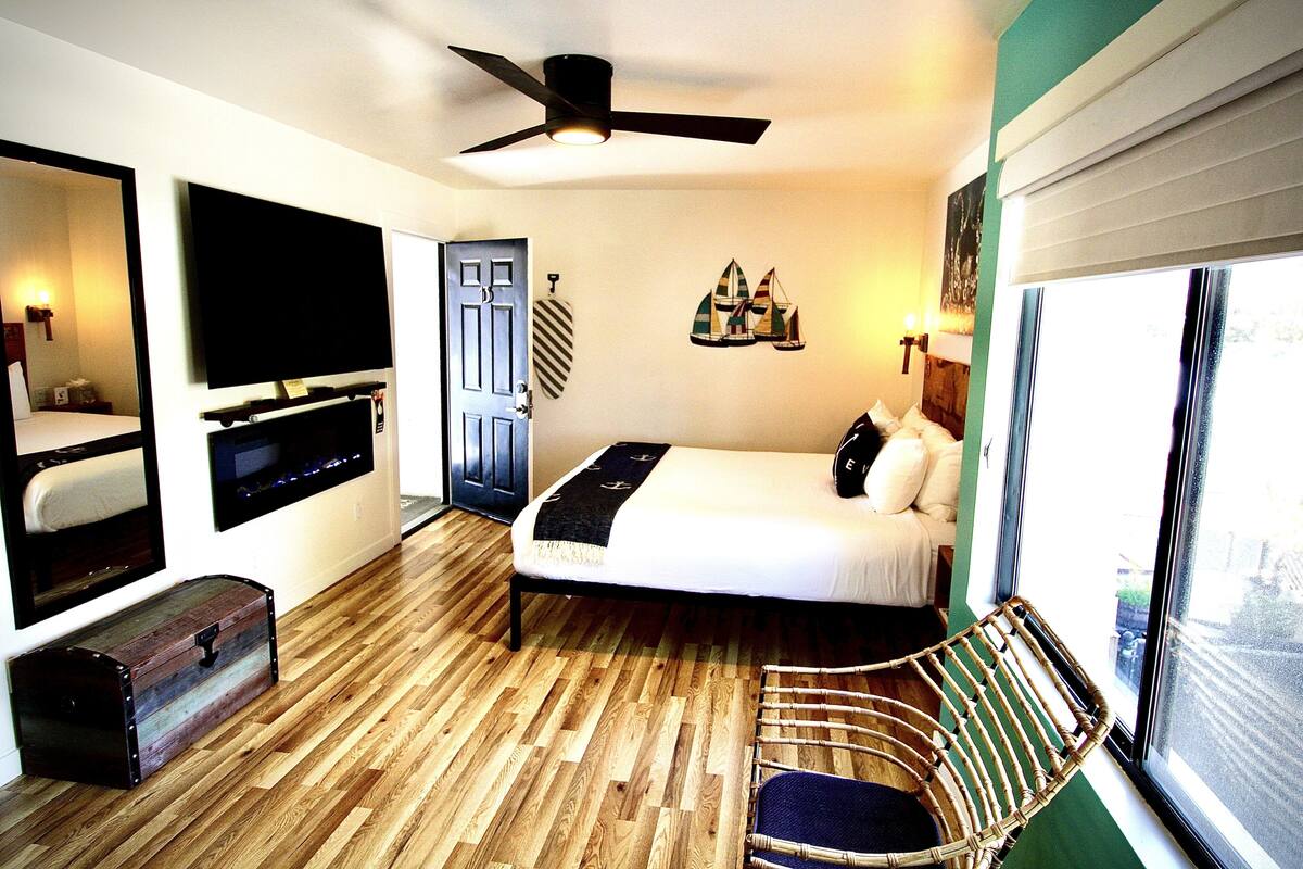 Quarto de hotel com cama, televisão, ventoinha no teto, janela e chão de madeira.