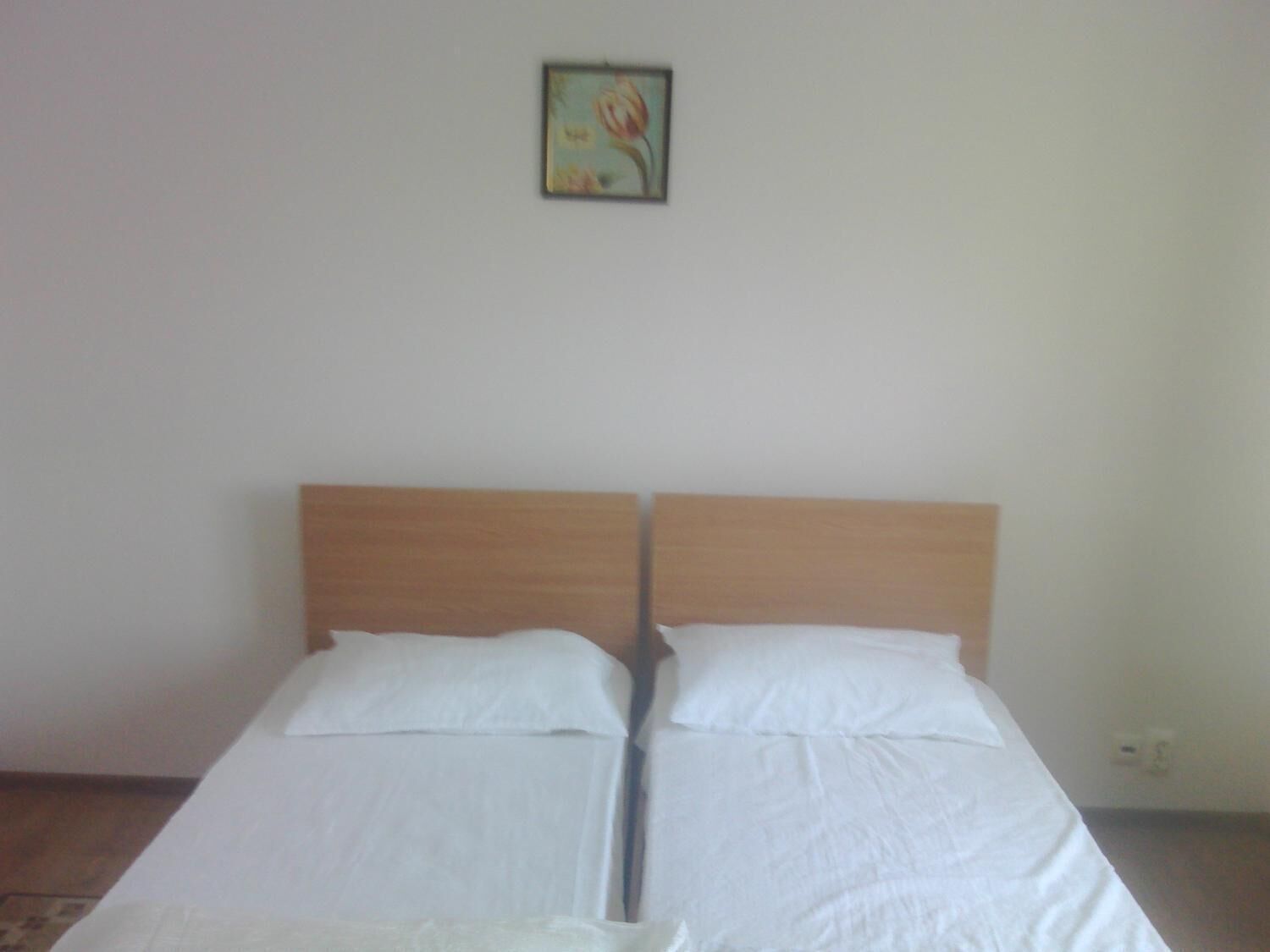 Quarto simples e limpo com cama de cabeceira em madeira, roupa de cama branca e um pequeno quadro na parede.