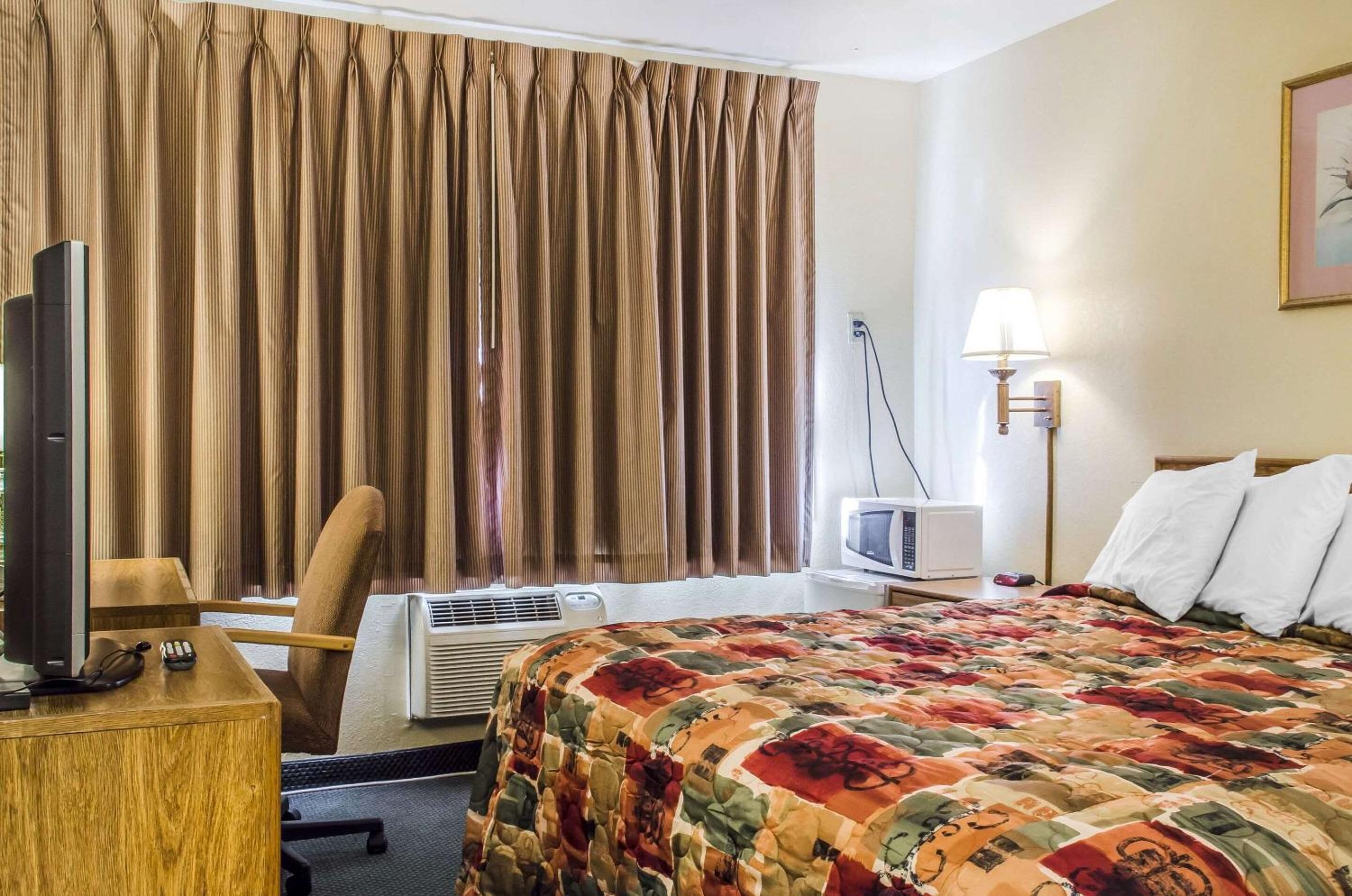 Quarto de hotel com cama, secretária com computador, cadeira, ar condicionado, televisão e luminária.